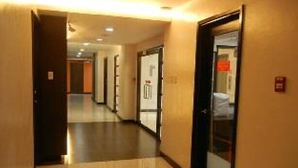 Um corredor de hotel com portas de acesso aos quartos, um alarme de incêndio na parede e um chão ladrilhado e limpo.