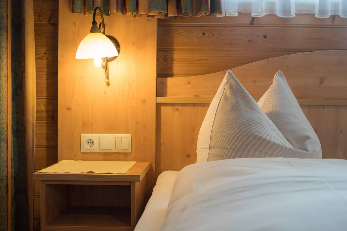 Uma cama de madeira com duas almofadas, uma mesinha de cabeceira e um abajur fixado na parede.