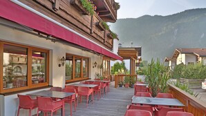 Um pátio externo com cadeiras e mesas vermelhas, deck de madeira e montanhas ao fundo.
