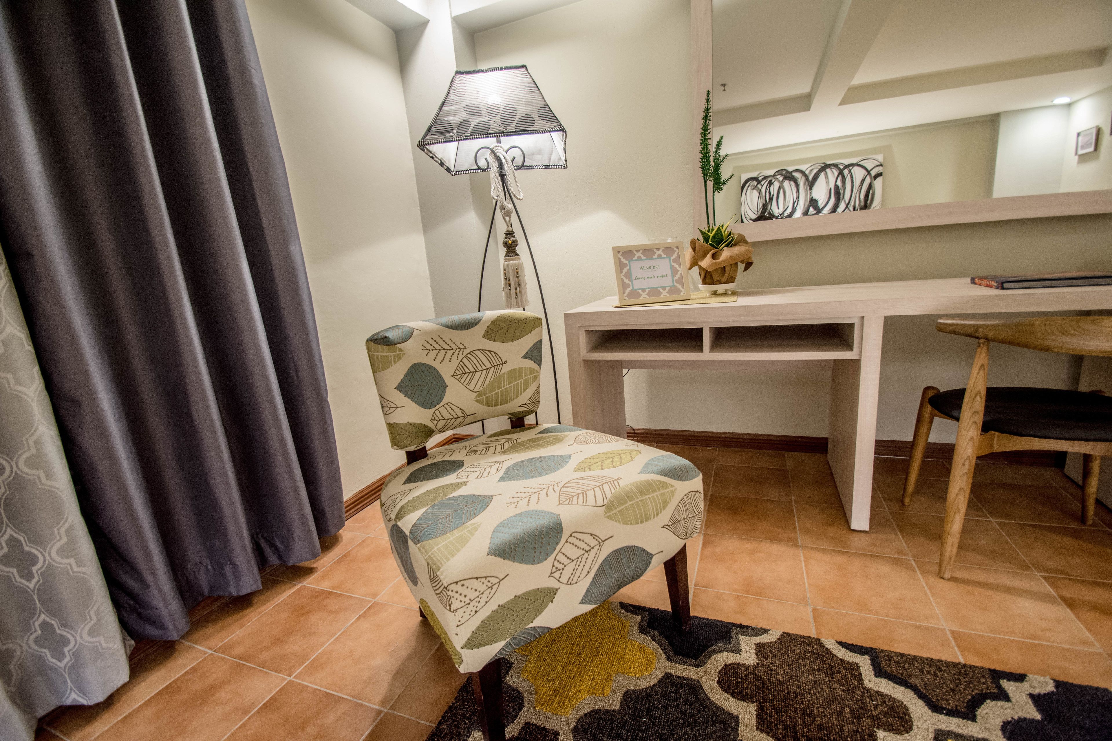 Uma sala de estar com uma poltrona estampada, uma escrivaninha, uma luminária e uma pequena planta.