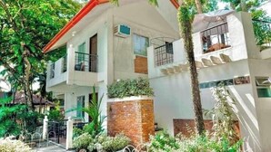 Um edifício de dois andares com telhado vermelho, paredes brancas e um detalhe em parede de tijolos. Possui varandas no andar superior e um pequeno espaço de pátio com plantas e uma mesa.