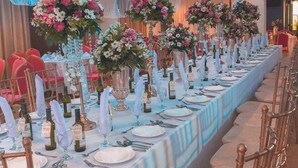 Uma longa mesa de banquete posta com toalhas brancas, arranjos florais e garrafas de vinho.