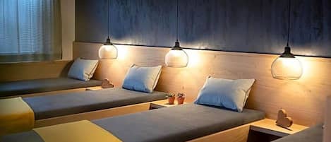 Quarto com três camas alinhadas na parede, cada uma com mesa de cabeceira e luminária.