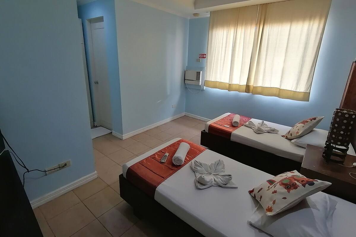 Quarto de hotel com duas camas, uma janela com cortinas, ar condicionado fixado na parede e uma porta.