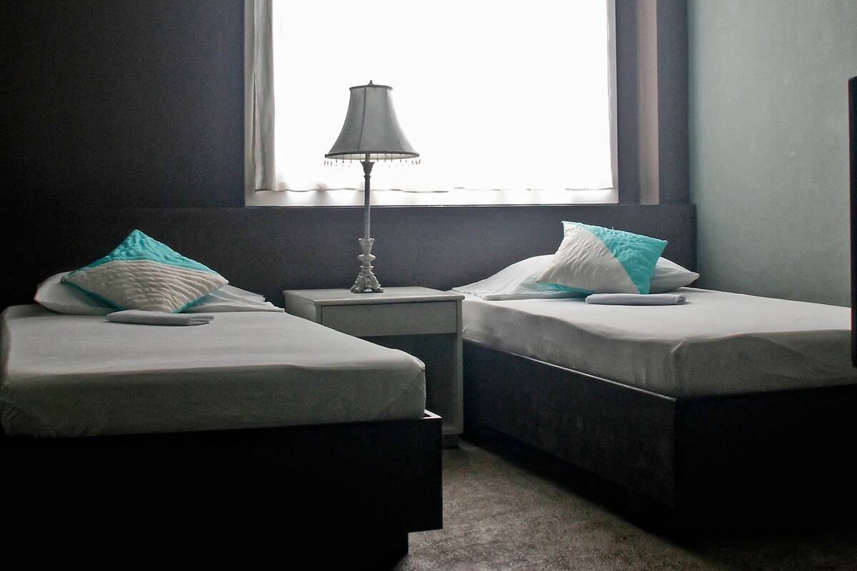 Quarto de hotel com duas camas de solteiro, uma mesa de cabeceira, uma luminária e uma janela com cortinas.