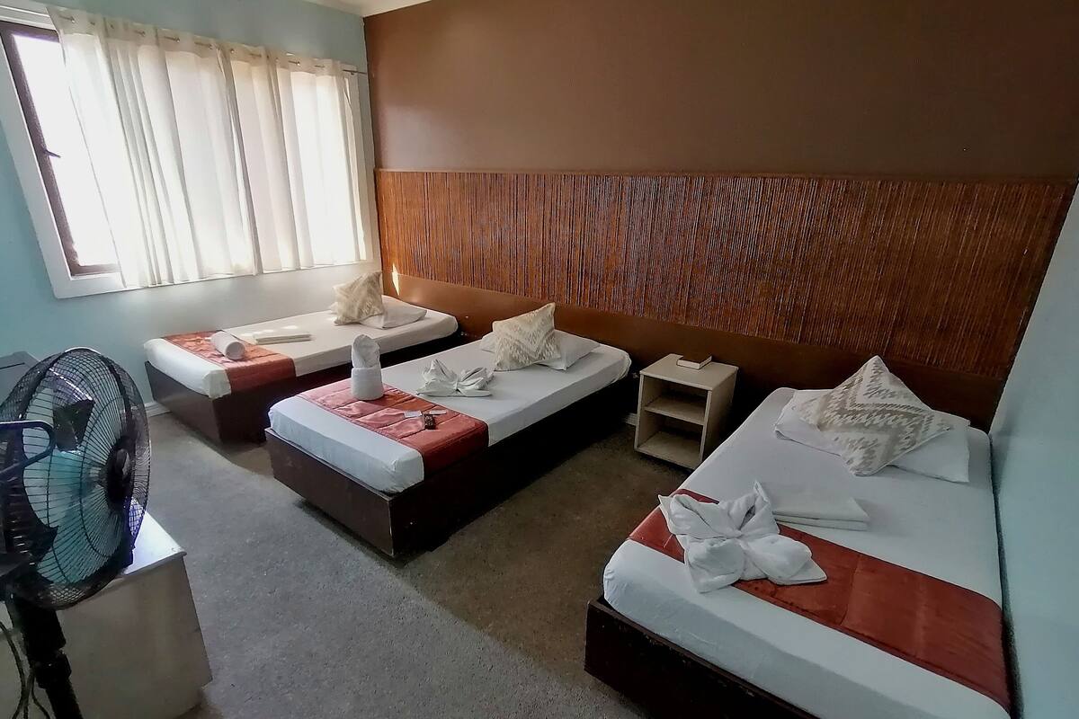 Quarto de hotel com três camas de solteiro, uma mesa de cabeceira pequena, um ventilador e uma janela com cortinas.