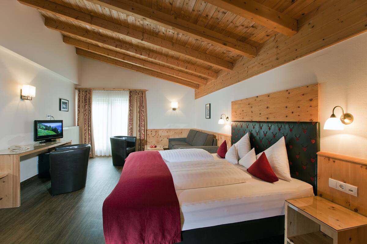 Quarto de hotel com teto de madeira, cama com roupa de cama vermelha e branca, escrivaninha com televisão e janela com cortinas.