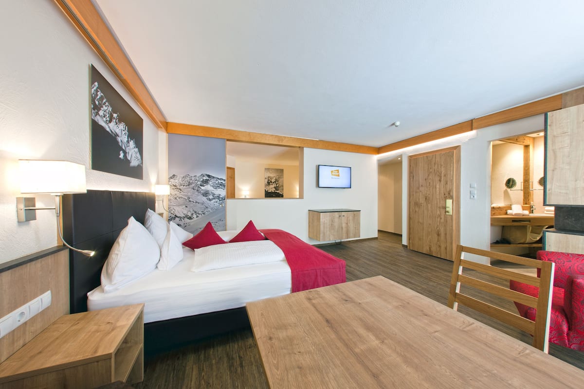 Quarto de hotel moderno com cama, mesas de cabeceira, escrivaninha e cadeira. Possui painel de cabeceira em madeira, um quadro pendurado e televisão na parede.