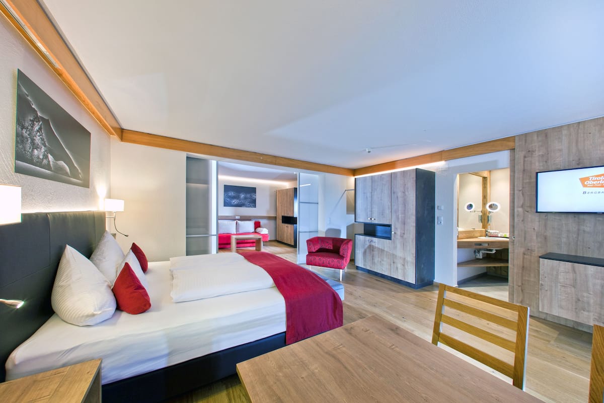 Quarto de hotel moderno com uma cama grande, uma mesa de jantar de madeira, uma poltrona vermelha e uma TV fixada na parede.
