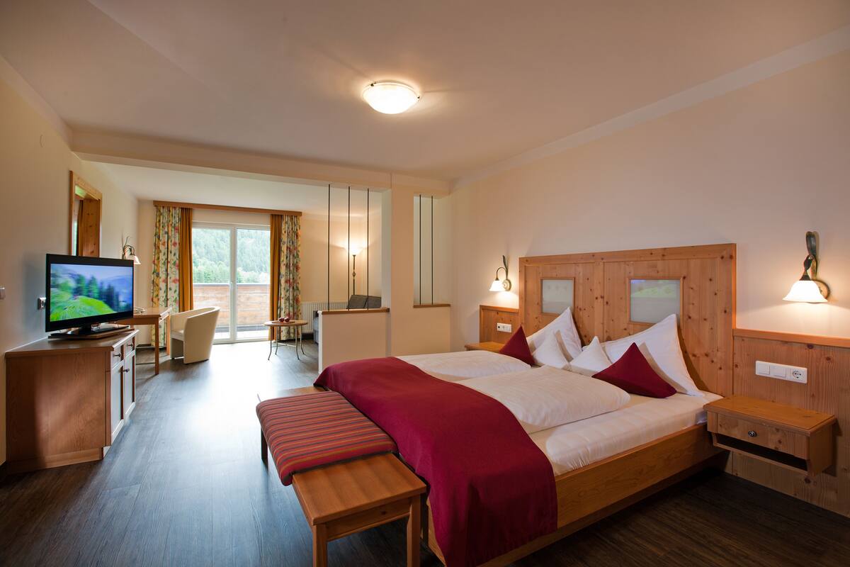 Quarto de hotel com uma cama grande, uma mesa com televisão, um banco e vista para o exterior através de uma janela.