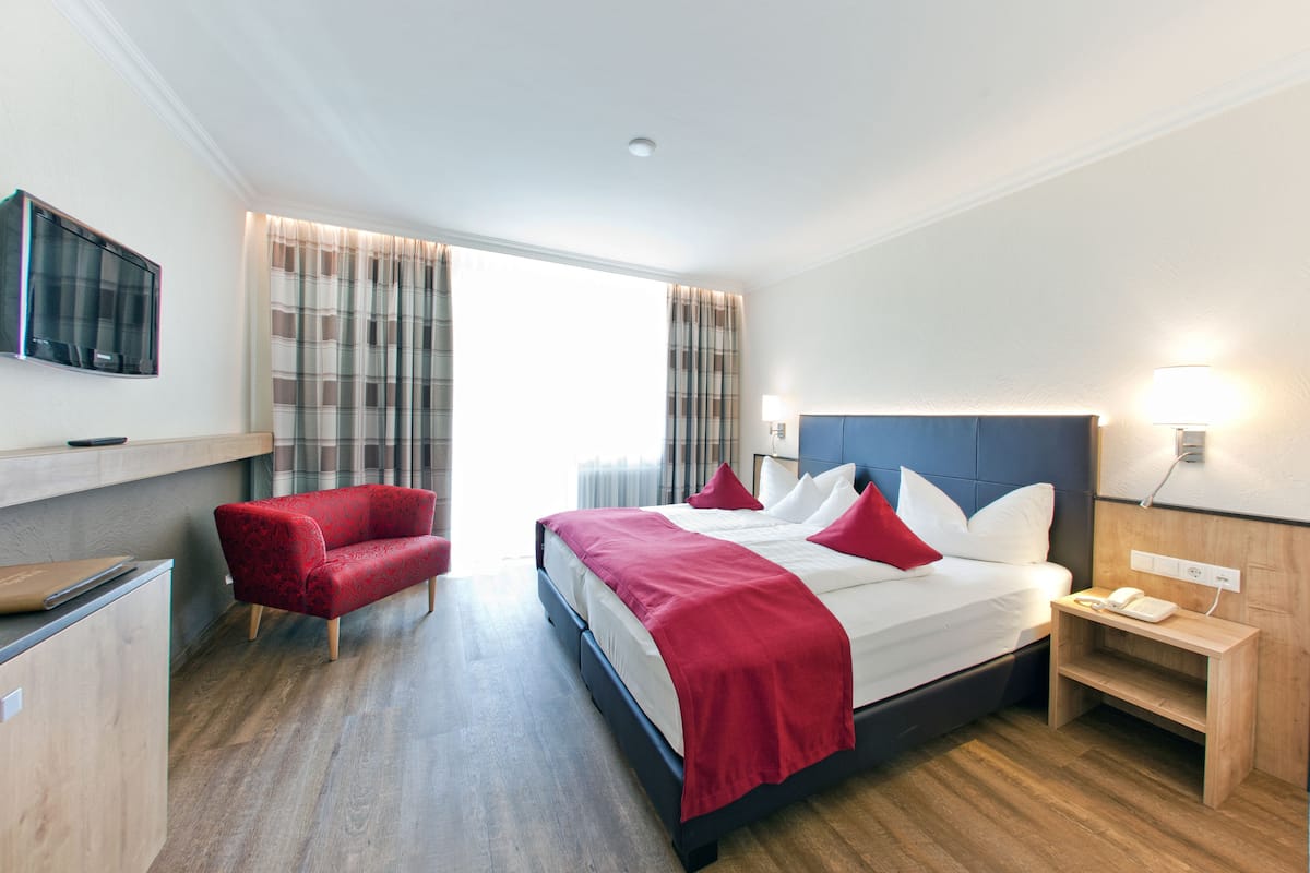 Quarto de hotel com uma cama, uma escrivaninha, uma cadeira, uma TV e uma janela com cortinas.