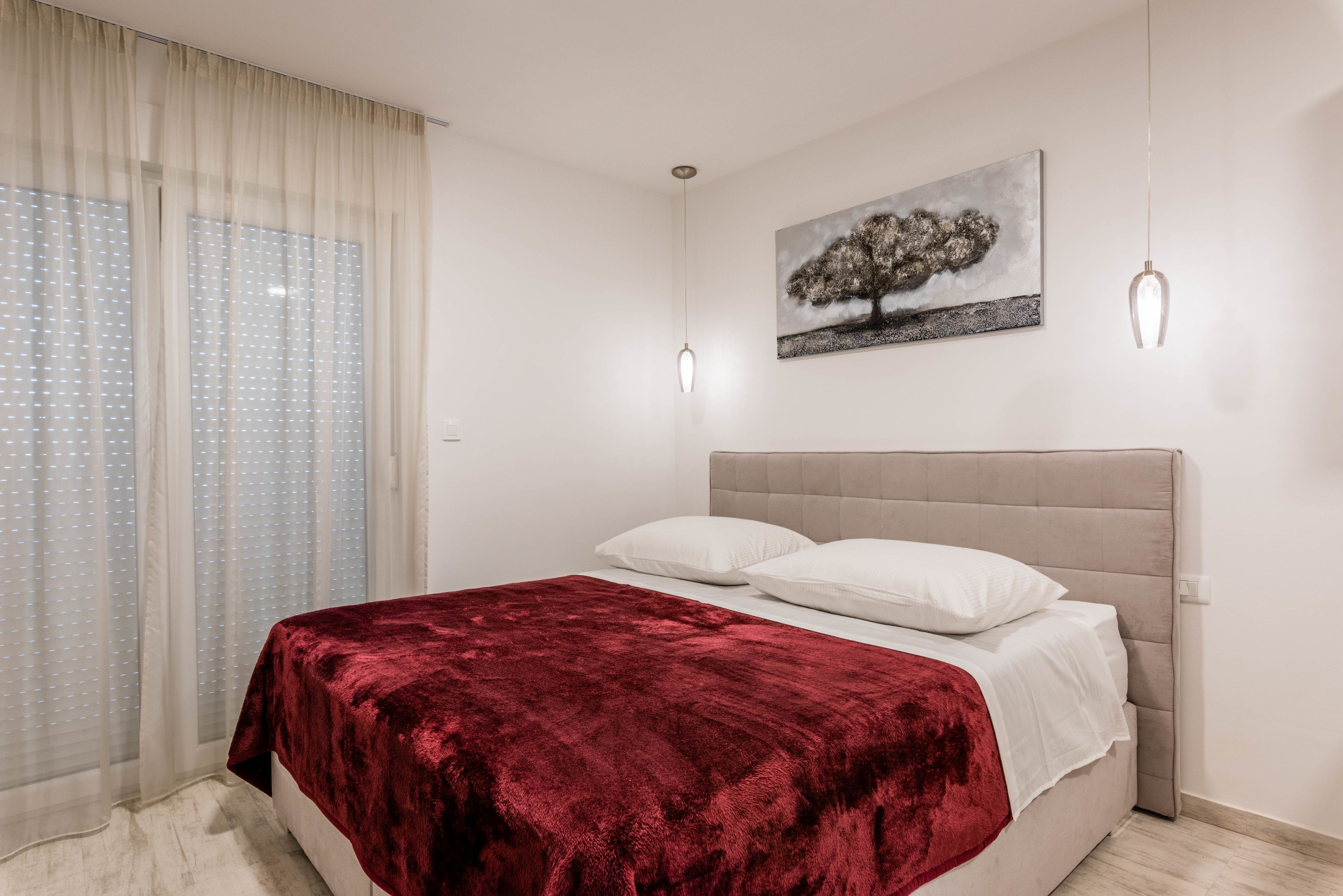 Quarto com cama, janela com cortinas leves, quadro pendurado na parede e duas luminárias pendentes.