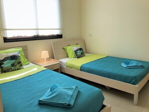 Quarto duplo com duas camas, cada uma com colcha verde e azul, mesa de cabeceira com abajur e janela com cortina branca.