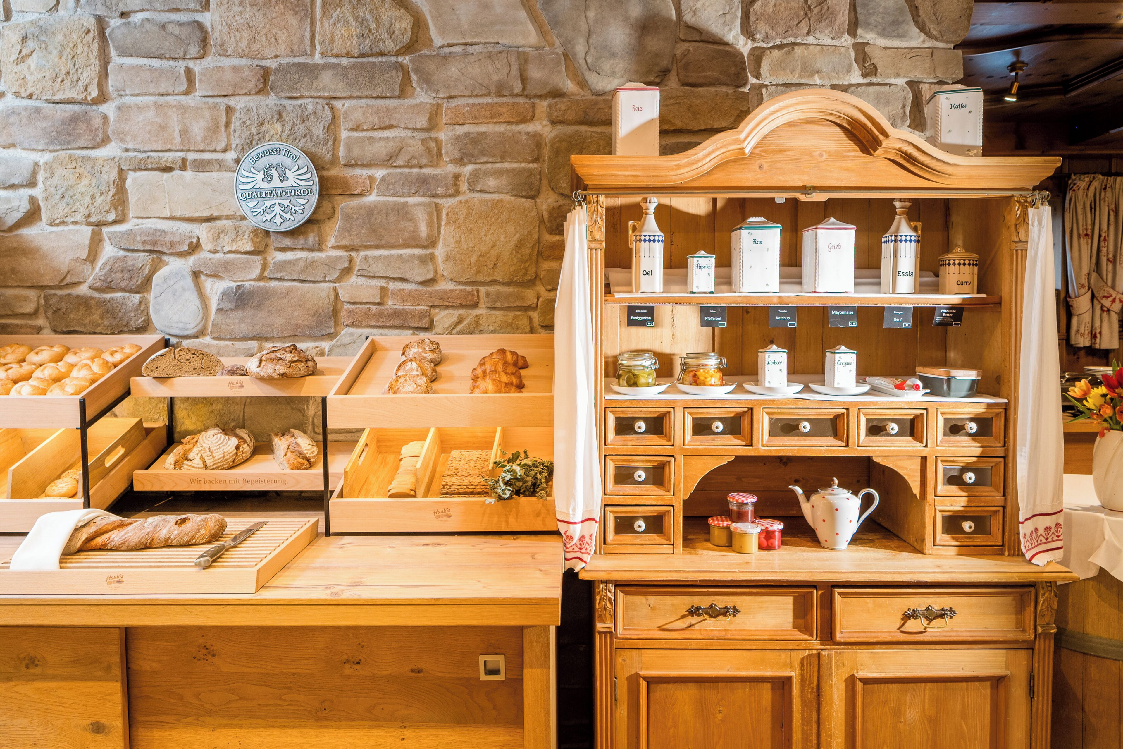 Vitrine de pastelaria com pão e doces, armários de madeira e parede de pedra ao fundo.