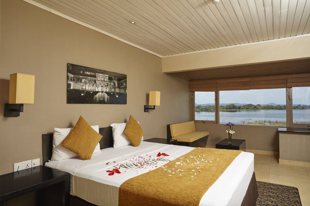 Quarto de hotel com cama, mesas de cabeceira, uma cadeira, escrivaninha, uma janela grande com vista para a água e um quadro na parede.