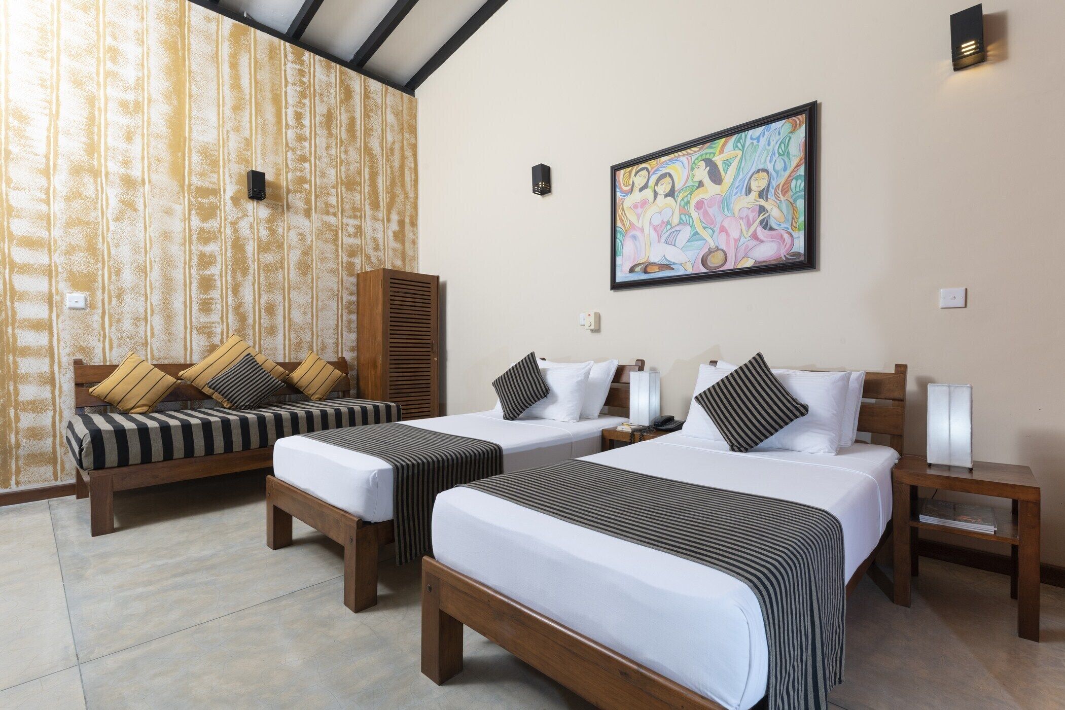 Quarto de hotel com duas camas de solteiro, um sofá-cama e uma cabeceira de madeira. Há um quadro na parede e uma pequena mesa com uma luminária.
