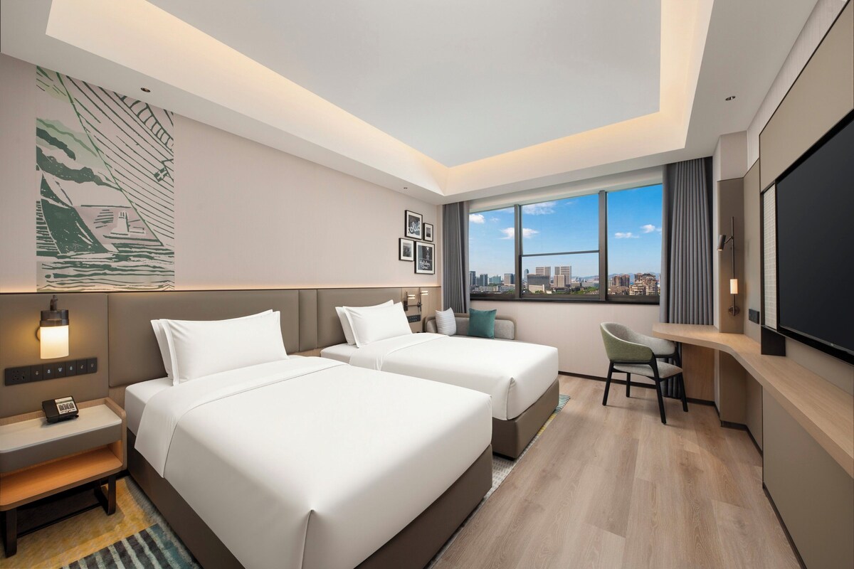 Quarto de hotel com duas camas, uma escrivaninha, uma cadeira, televisão e uma janela ampla com vista para a cidade.