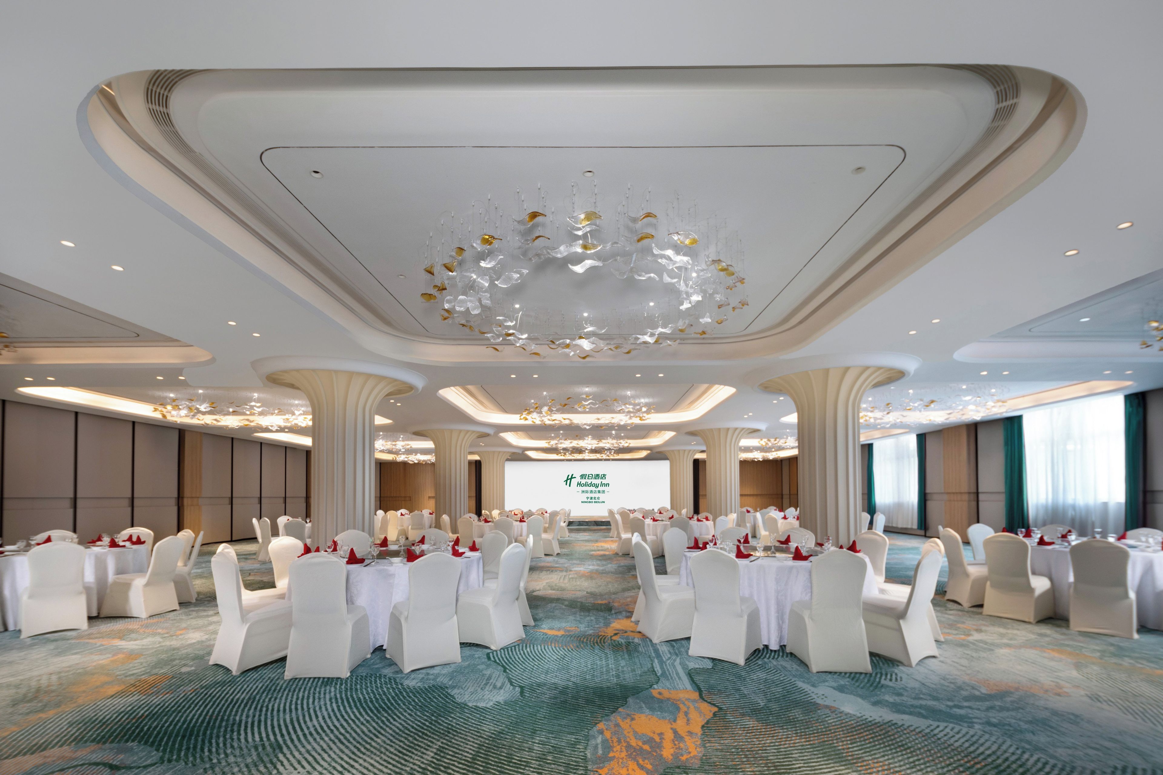Uma grande sala de conferências com cadeiras brancas, palco central e um lustre moderno.