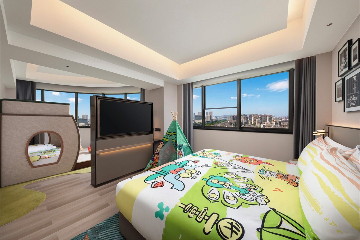 Quarto de hotel moderno com uma cama grande, televisão, um espaço de recreação pequeno e vista para a cidade.