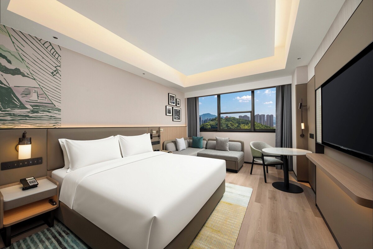 Quarto de hotel moderno com uma cama grande, um sofá, uma mesinha e uma televisão.