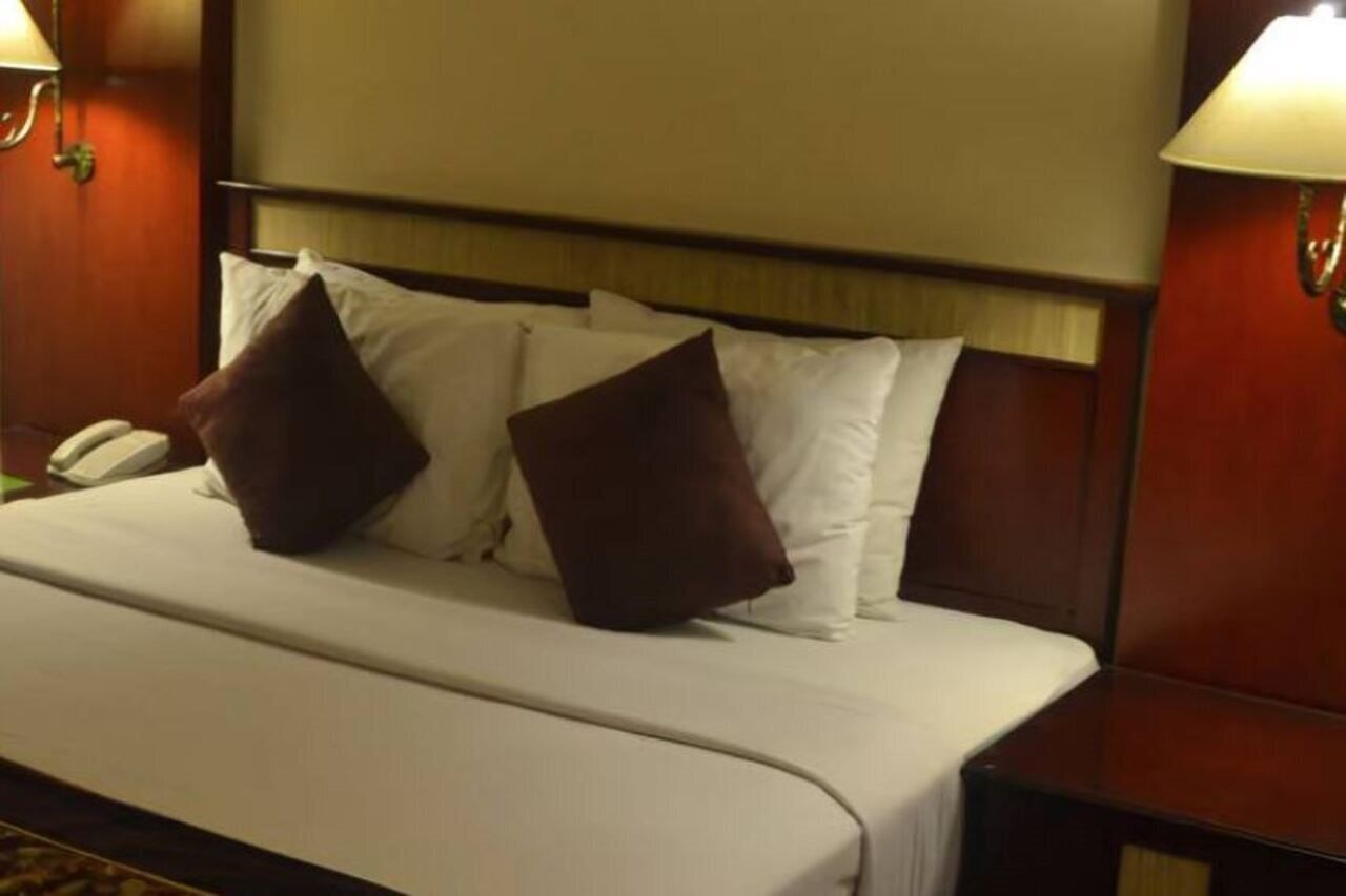 Quarto de hotel com cama, almofadas, mesa de cabeceira e abajur.