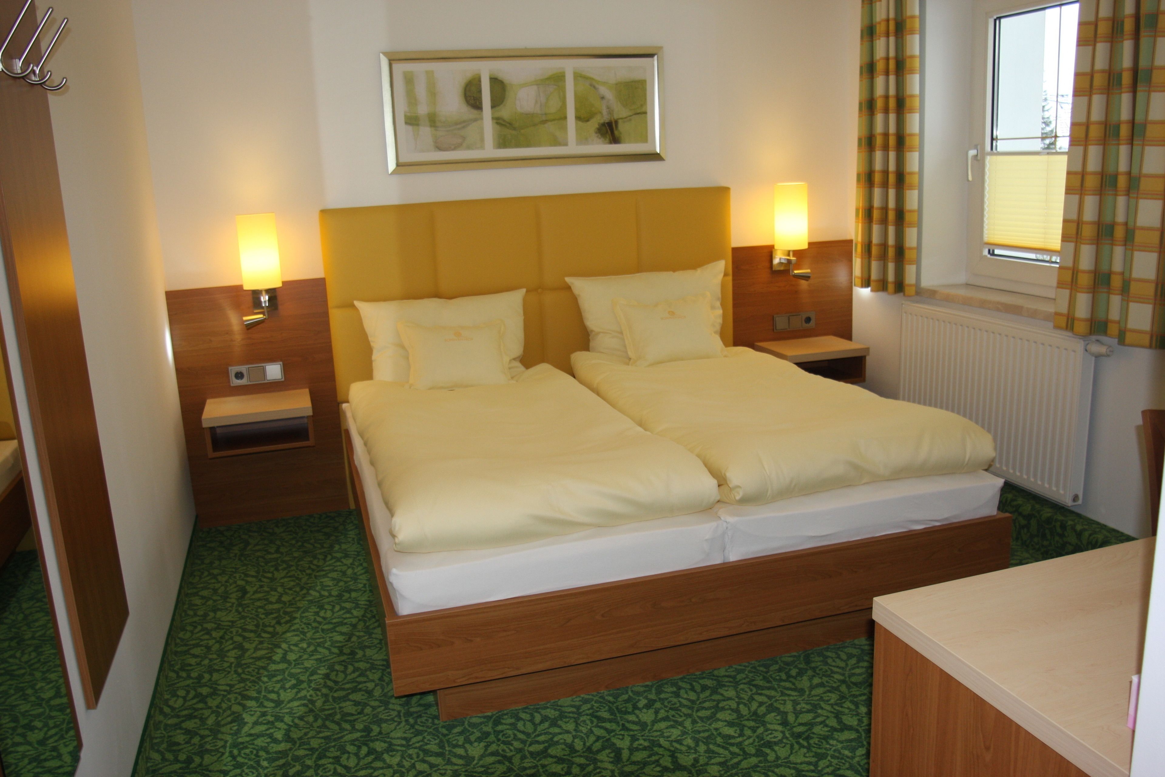 Cama de casal com cabeceira de madeira, duas mesas de cabeceira com abajures, um quadro emoldurado acima da cama e uma janela com cortinas.