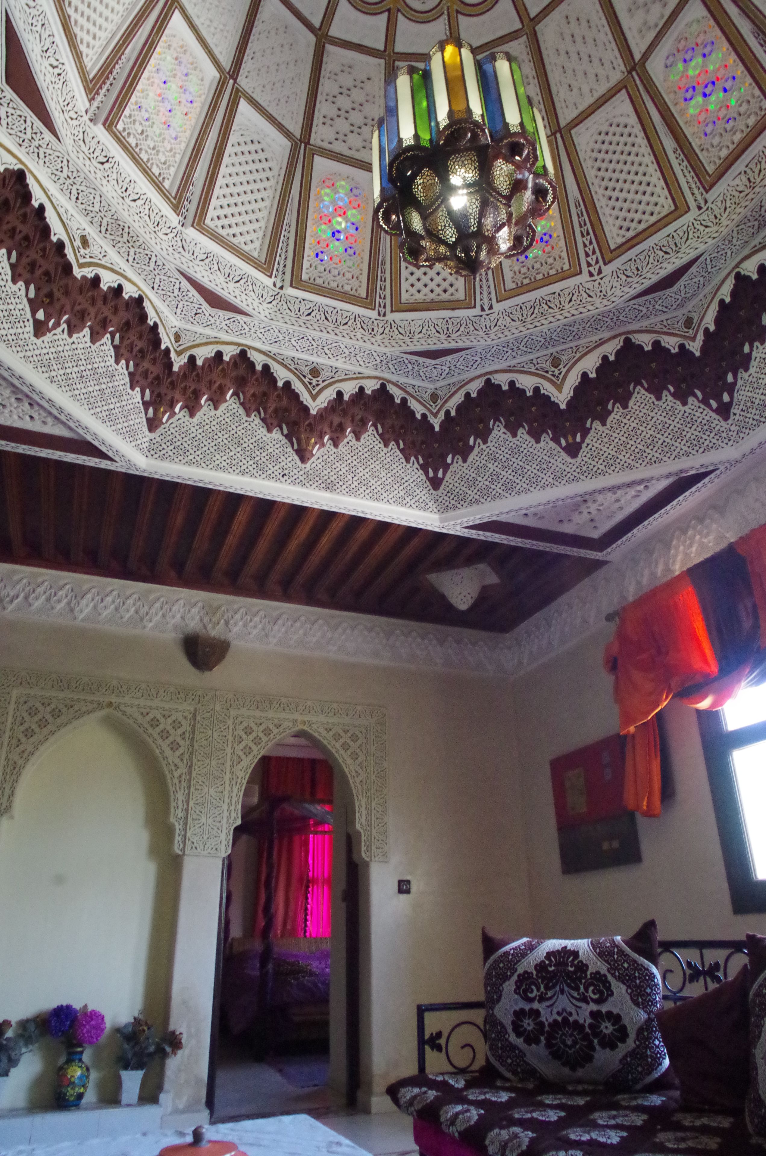 Quarto com padrões intrincados no teto, cama com almofadas estampadas e uma porta que dá para outro cômodo.