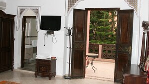 Quarto com televisão fixada na parede, um armário de madeira e uma grande porta de madeira que dá para área externa com vista para vegetação.