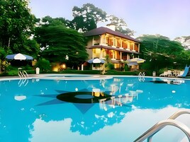 Piscina com superfície de água azul cristalina, rodeada por vegetação exuberante e um edifício grande com várias varandas.