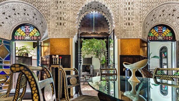 Interior com arcos decorados, uma mesa de vidro e cadeiras estampadas.