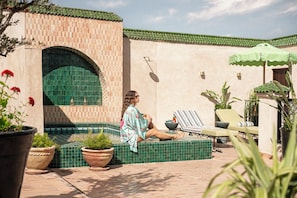 Uma pessoa relaxando perto de uma piscina com parede azulejada, luminária pendente e plantas em vasos.