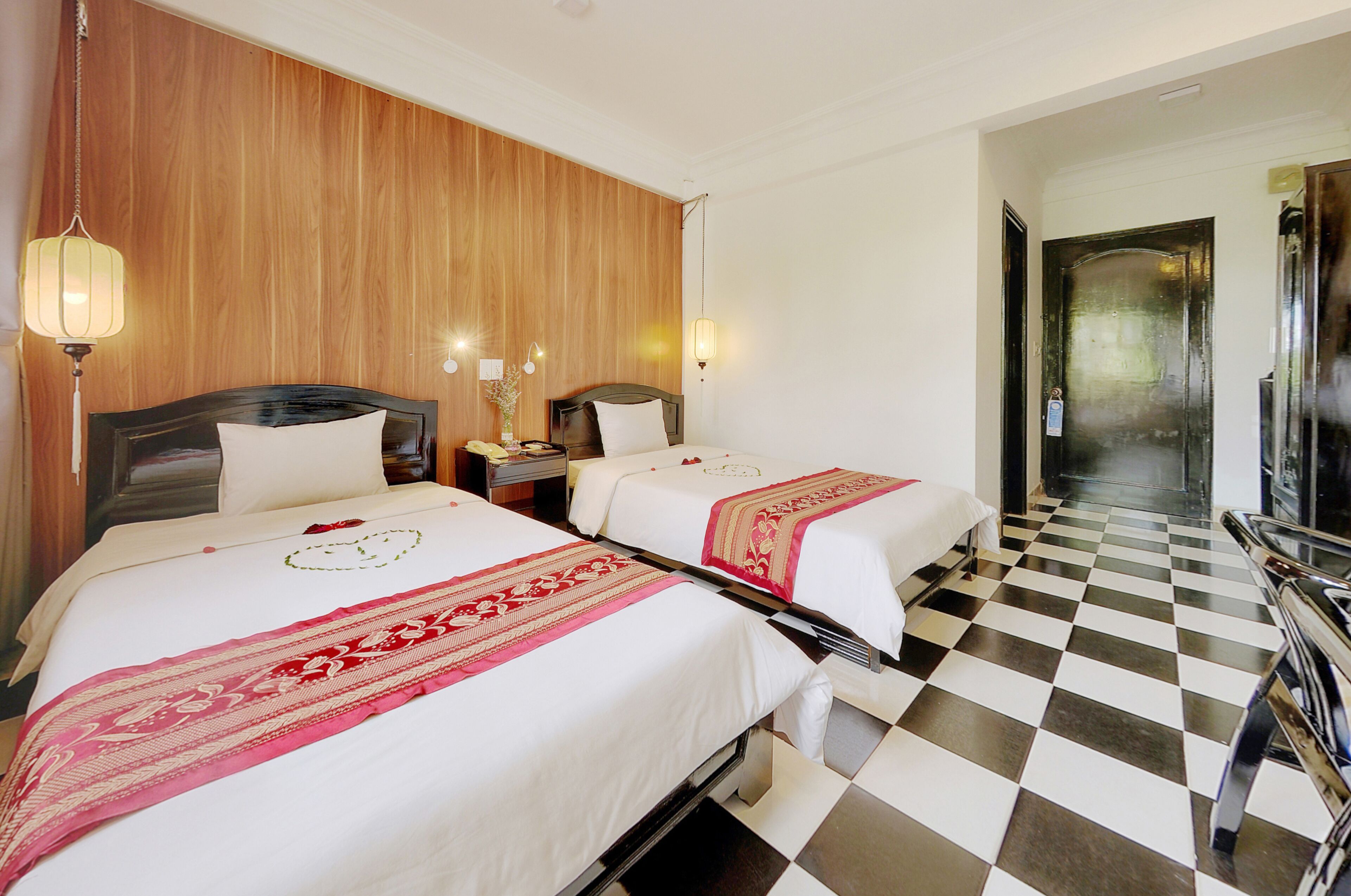 Quarto de hotel com duas camas, piso quadriculado, painéis de madeira na parede e uma porta visível.