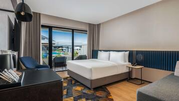 Quarto de hotel com uma cama, escrivaninha, cadeira, mesa pequena, luminária e vista para uma piscina e edifícios.