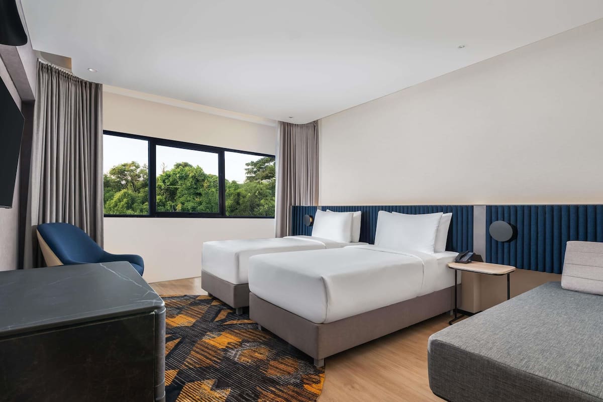 Quarto de hotel com duas camas, uma escrivaninha, uma cadeira, uma televisão e uma janela com vista para o verde.