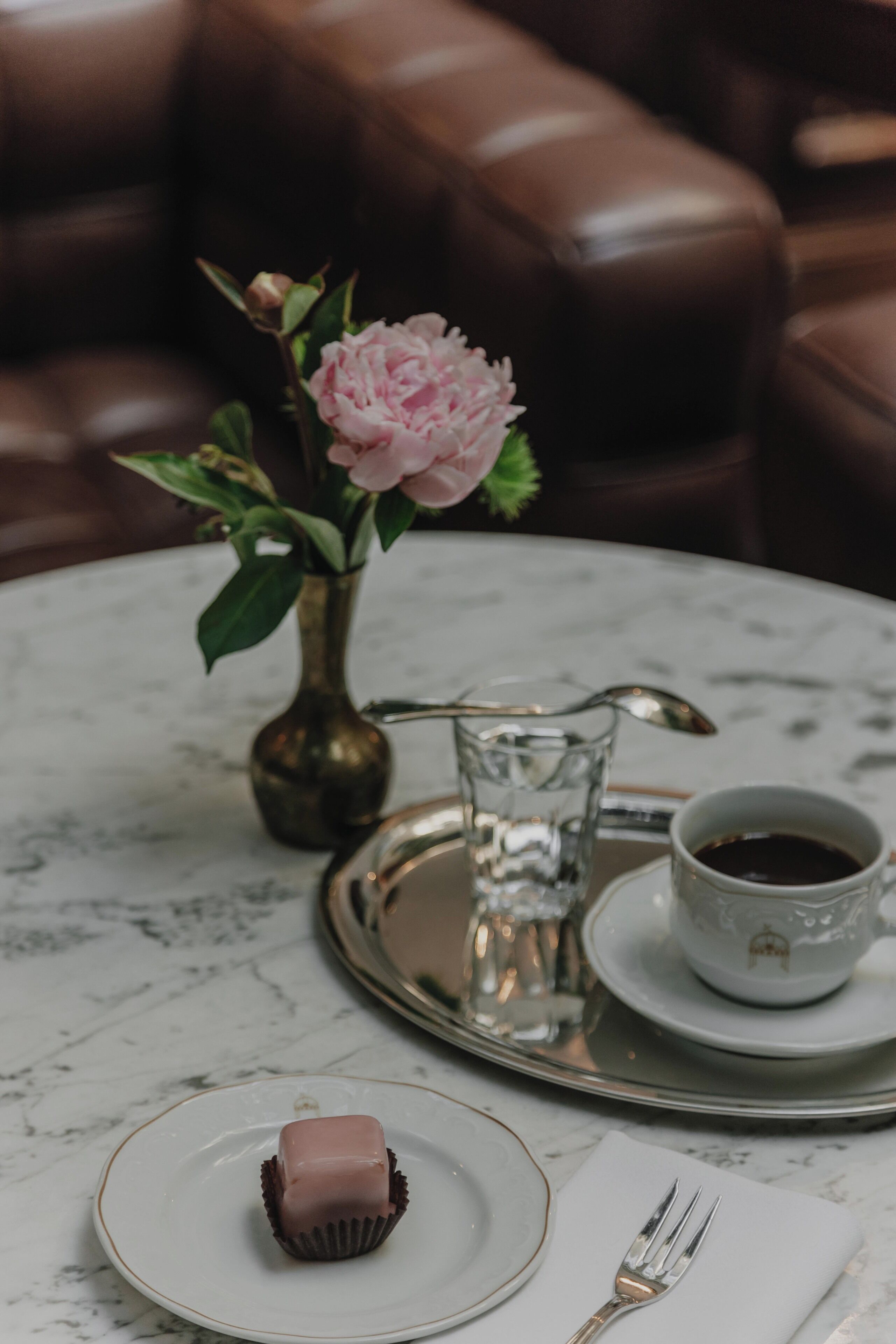 Uma mesa redonda com um prato branco, um copo d'água, uma xícara de café, um pequeno vaso com uma flor rosa e um garfo sobre um guardanapo.