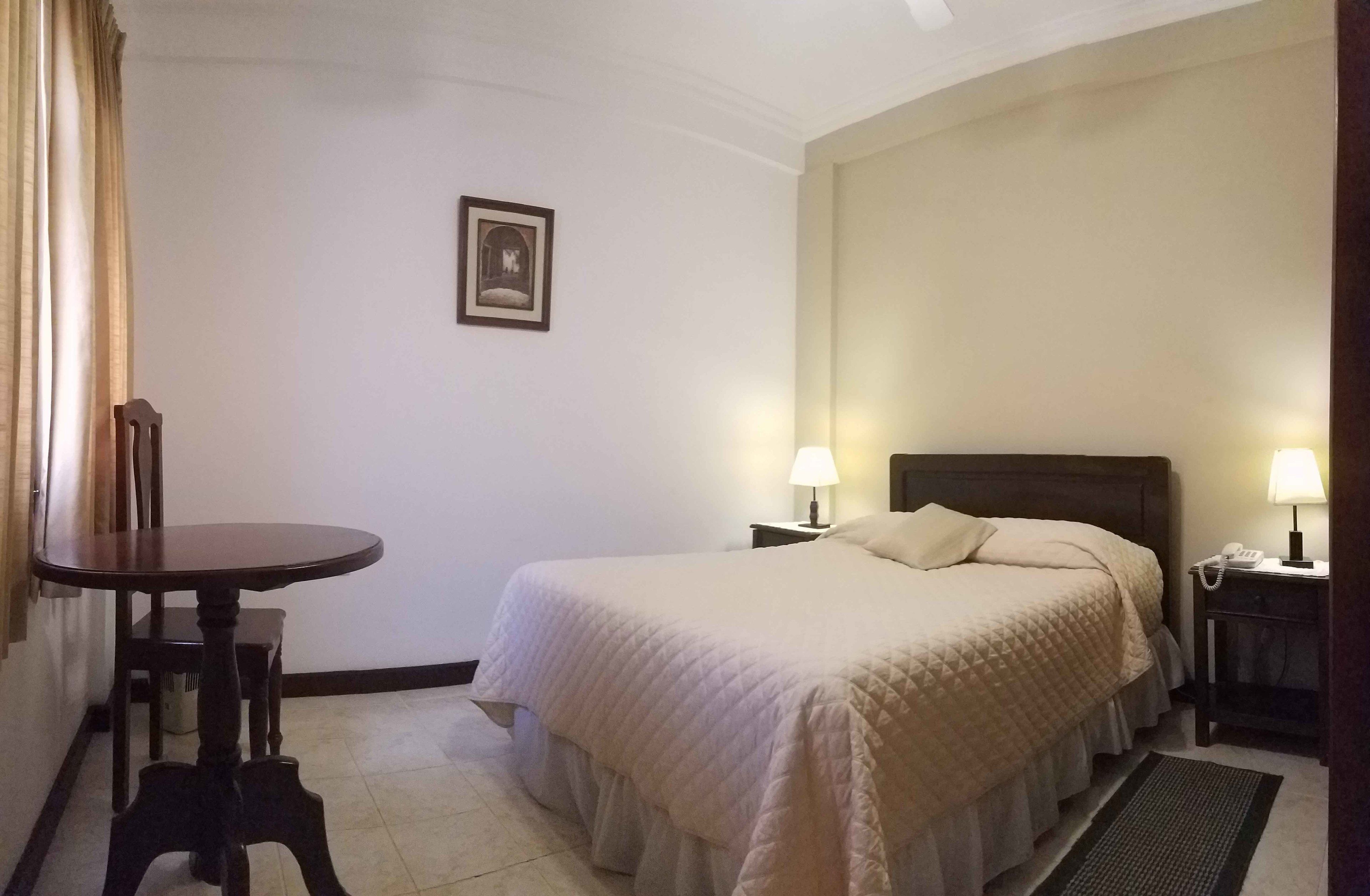 Quarto de hotel com uma cama, duas luminárias de cabeceira, uma pequena mesa com uma cadeira, um quadro na parede e uma janela com cortinas.