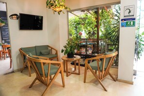 Quarto com sofá, duas cadeiras, uma mesinha, planta em vaso, televisão e janela com vista para o verde.