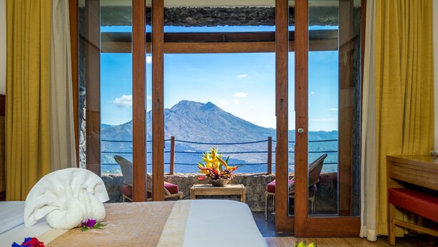 Quarto de hotel com cama, mesa com arranjo de flores e vista para uma montanha através de grandes portas de vidro.
