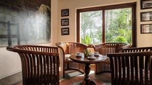 Uma área de refeições com cadeiras de madeira e uma mesa, uma janela grande com vista para o verde, e quadros na parede.