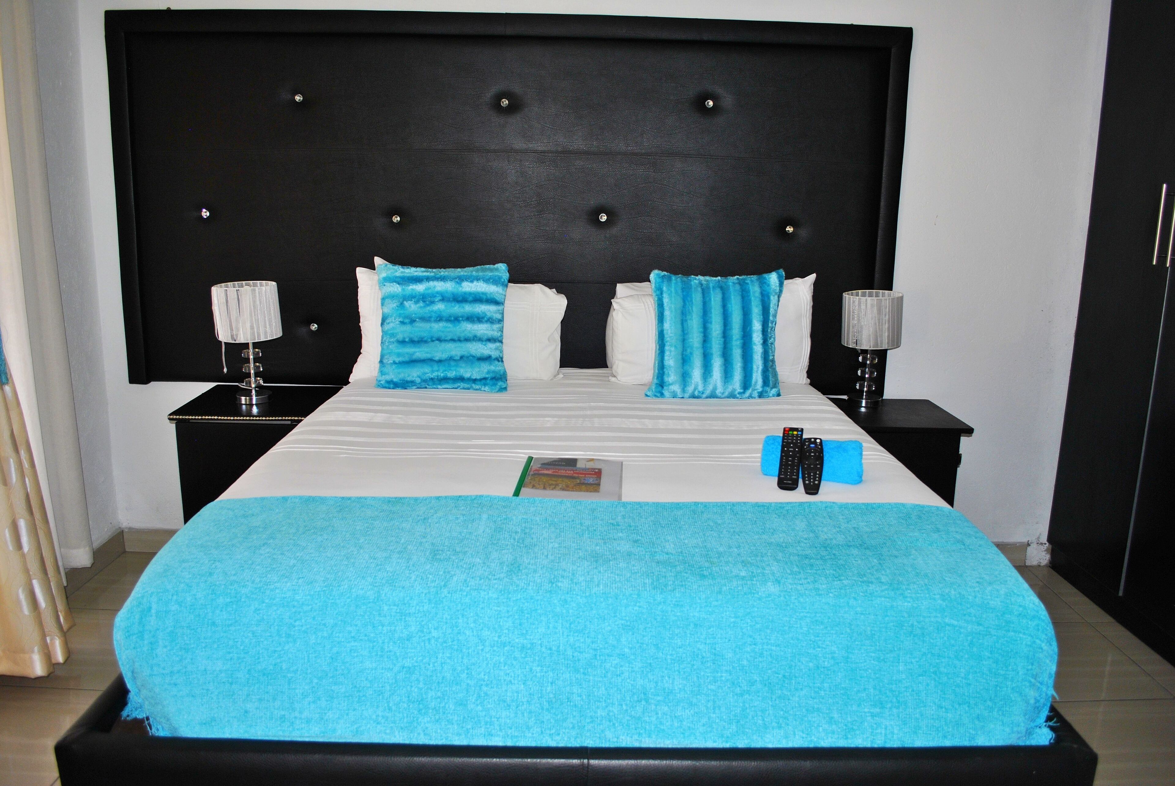 Uma cama bem arrumada com um cobertor azul, duas almofadas e um livro sobre a cama. Há duas mesas de cabeceira com luminárias e controles remotos.