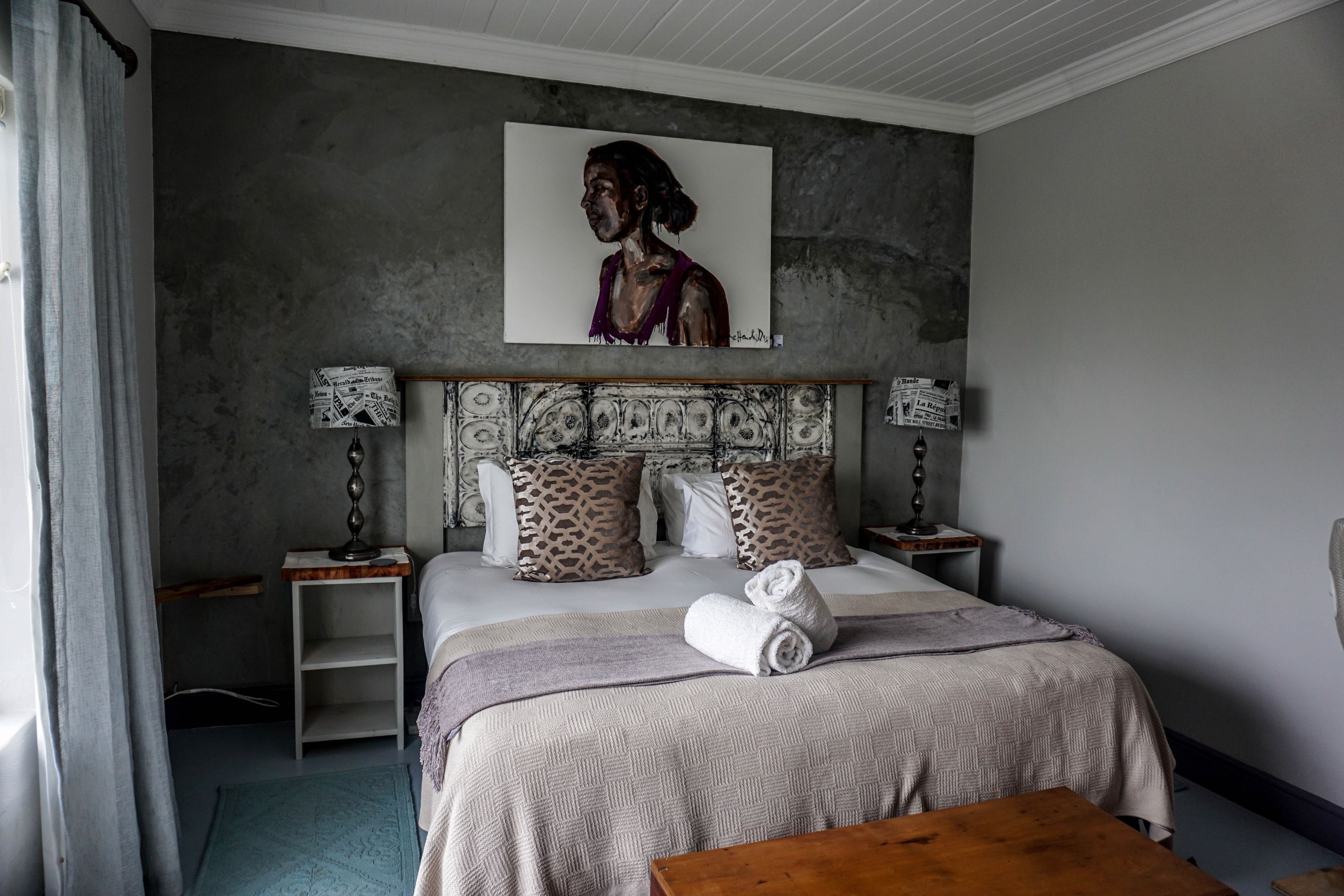 Quarto com cama grande, duas mesas de cabeceira, cabeceira com padrão decorativo, um quadro sobre a cama e janela com cortinas.