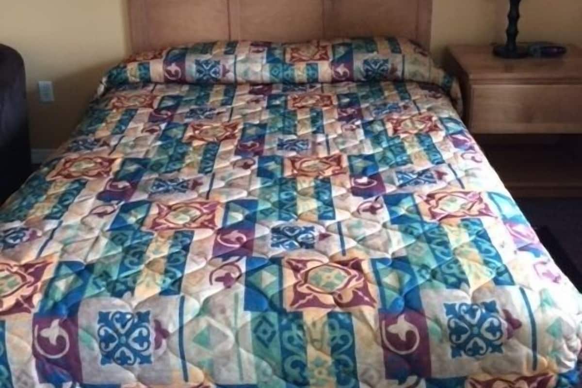 Uma cama com um colcha colorida, uma mesa de cabeceira com abajur e um quadro na parede.