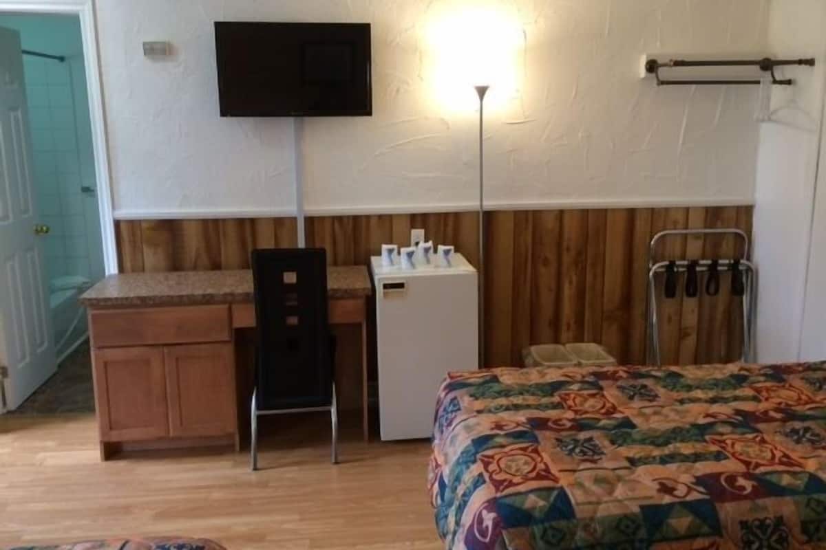 Quarto pequeno e compacto com escrivaninha de madeira, uma cadeira, cama com colcha colorida, frigorífico branco pequeno e televisão montada na parede.