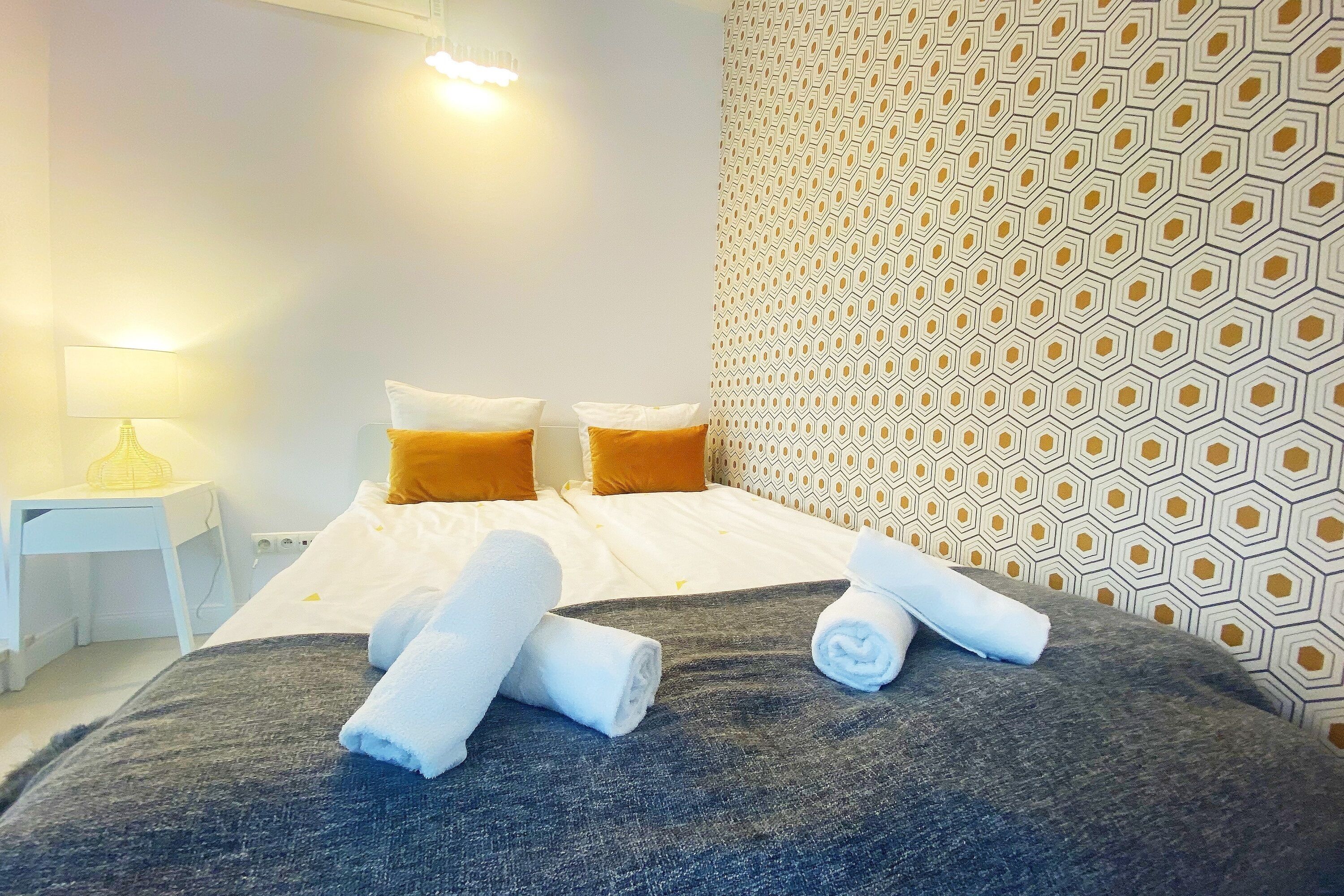 Quarto de hotel com cama, duas almofadas, mesa de cabeceira com abajur e parede estampada.