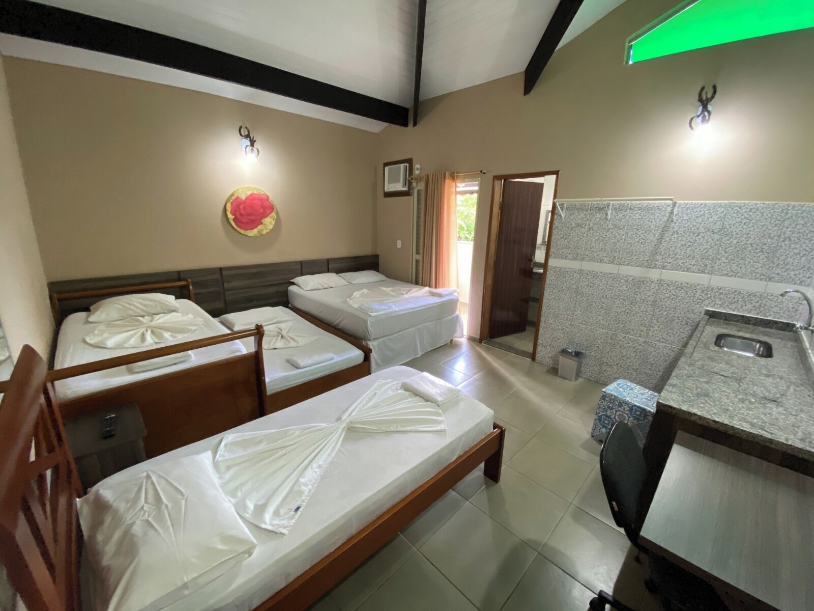 Quarto de hotel com quatro camas, banheiro com pia e espelho, e porta que dá para o exterior.