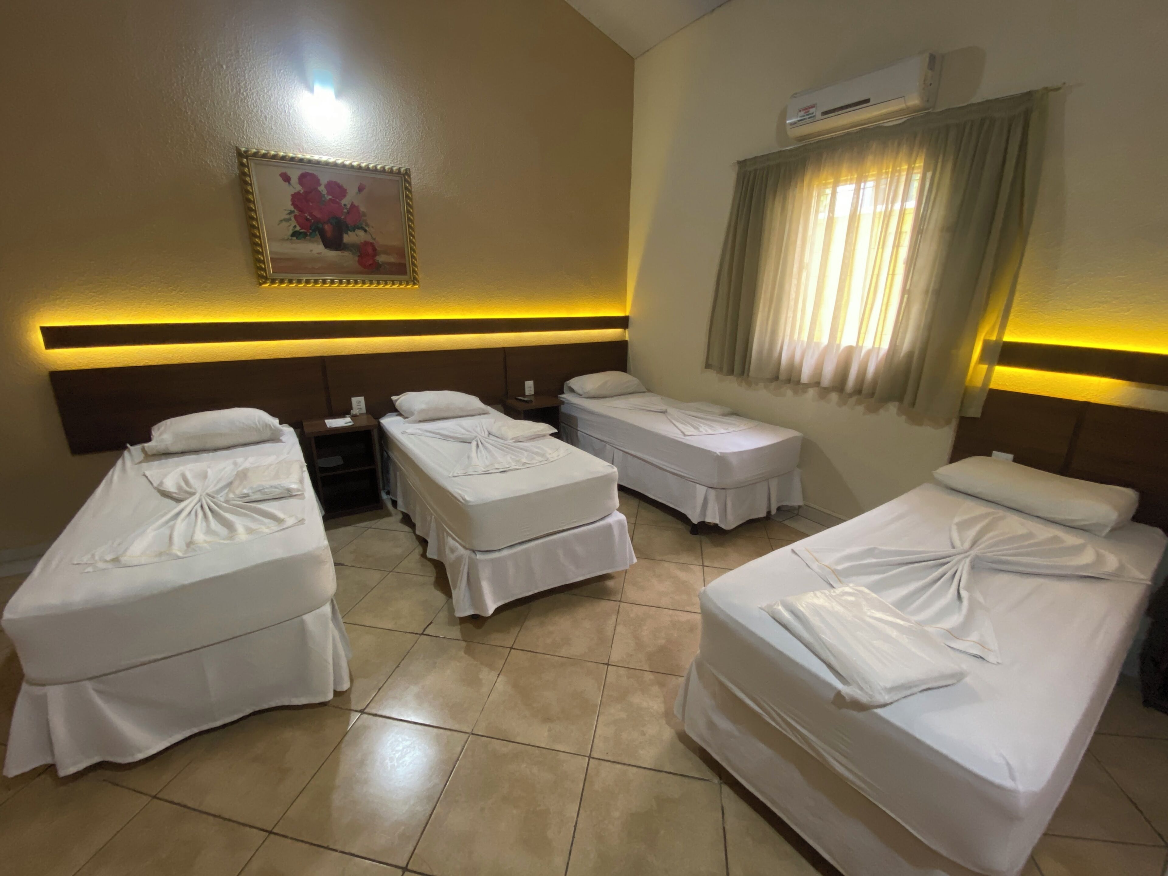 Quarto de hotel com quatro camas de solteiro, mesa de cabeceira com abajur, ar-condicionado fixado na parede e quadro pendurado acima das camas.