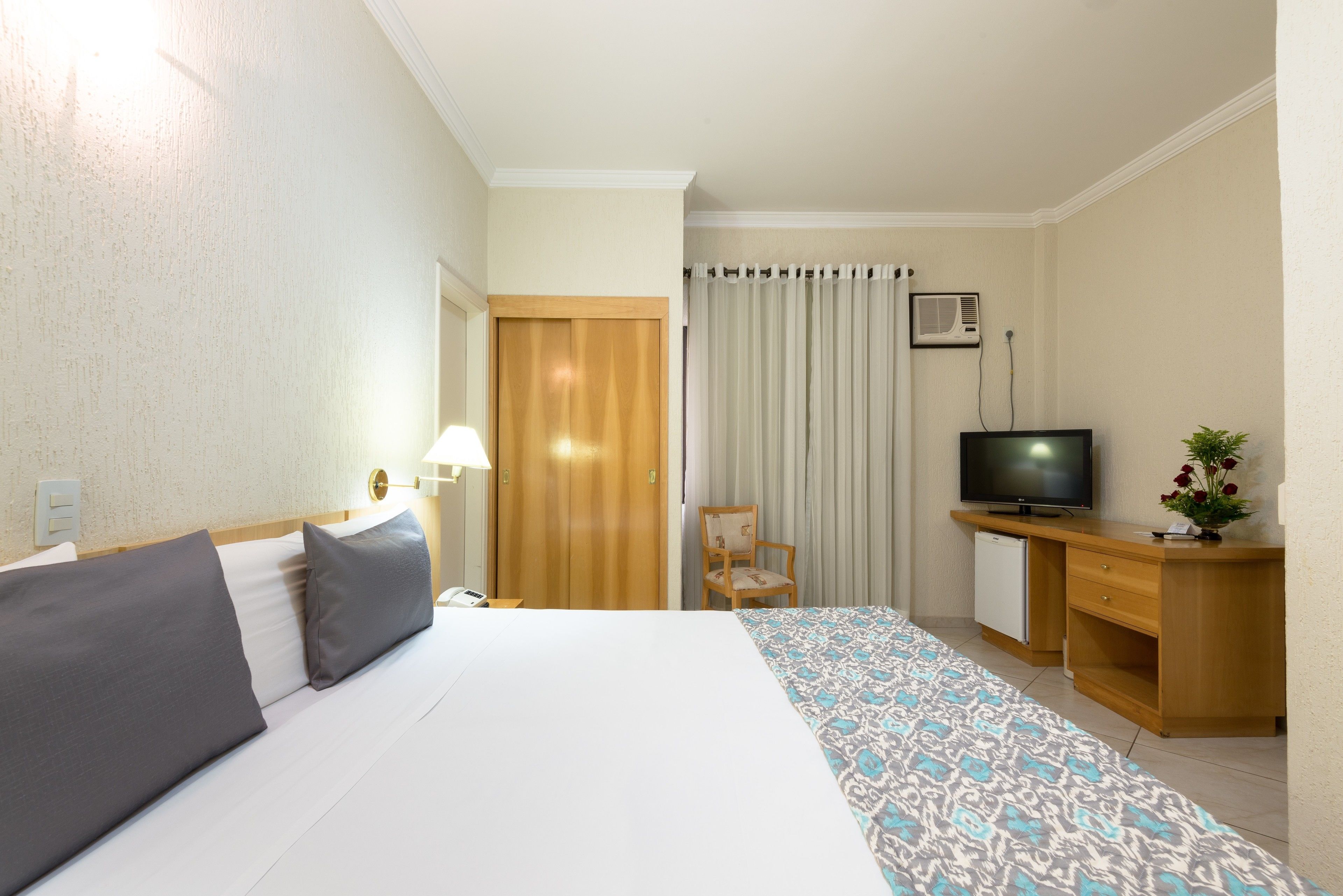 Quarto de hotel com uma cama grande, uma escrivaninha com televisão, uma cadeira e um pequeno frigorífico.