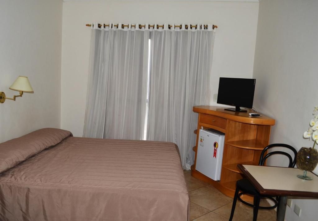 Quarto de hotel com cama, escrivaninha com computador, pequena geladeira e uma cadeira.