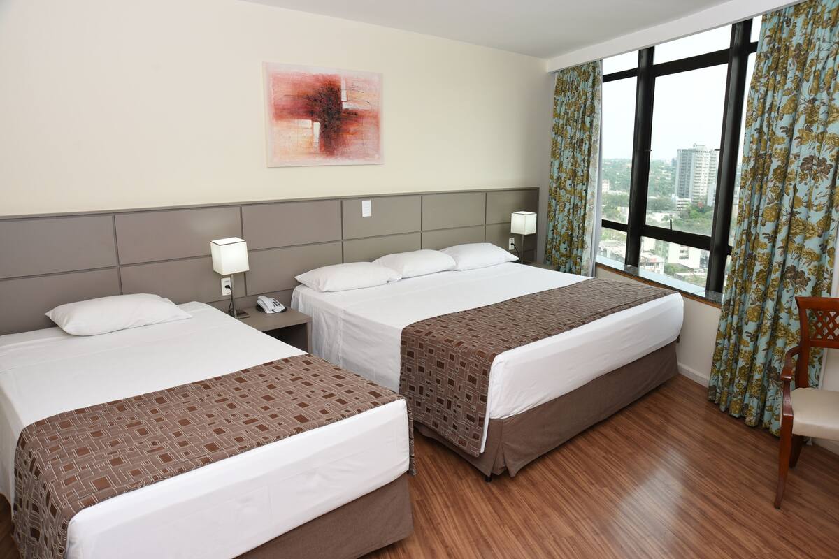 Quarto de hotel com duas camas, mesa de cabeceira com telefone, luminária na parede e janela com cortinas.