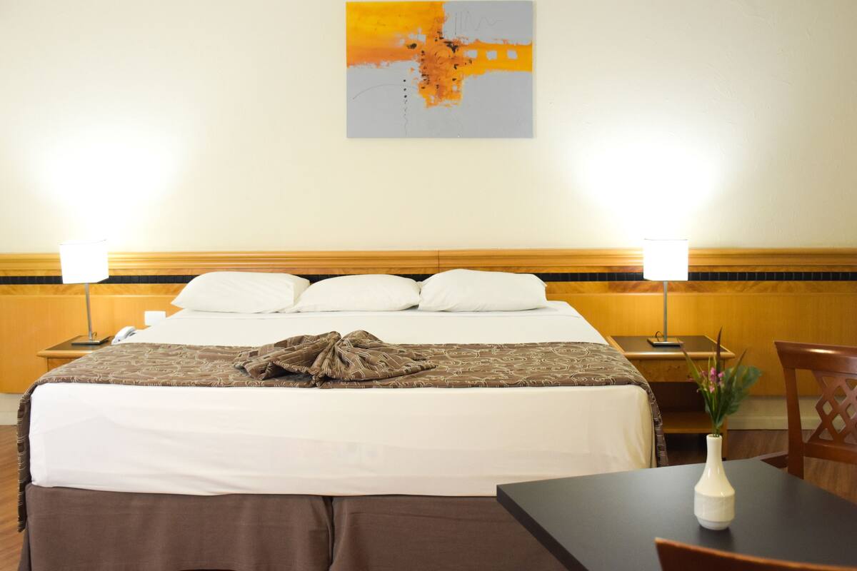 Quarto de hotel com cama, mesas de cabeceira, luminária, um vaso com flores e um quadro na parede.